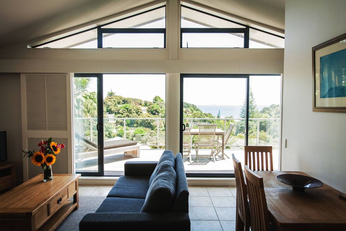 Uma sala de estar moderna com sofá, mesa de jantar e vista para uma varanda com mobiliário externo.
