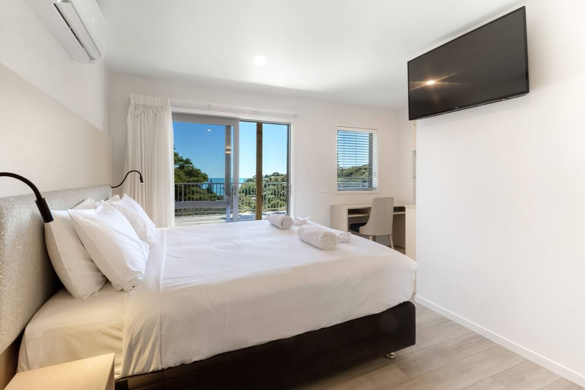 Quarto moderno com uma cama grande, TV fixada na parede e varanda com vista para o mar.