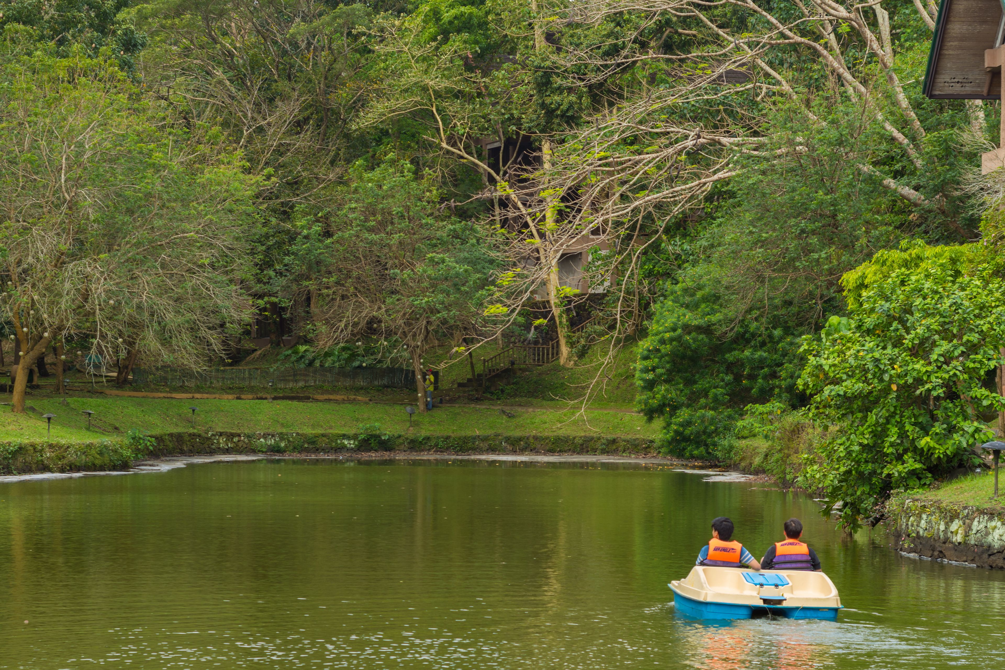 Um lago tranquilo com um barco transportando duas pessoas, cercado por vegetação exuberante e um edifício de pedra ao fundo.
