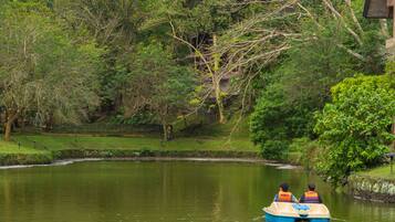 Um lago tranquilo com um barco transportando duas pessoas, cercado por vegetação exuberante e um edifício de pedra ao fundo.