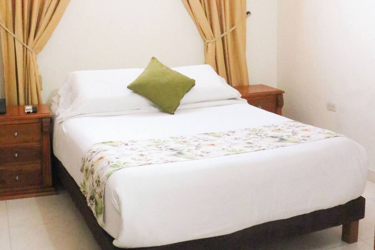 Uma cama bem arrumada com colcha floral, duas mesas de cabeceira e um ar condicionado fixado na parede.