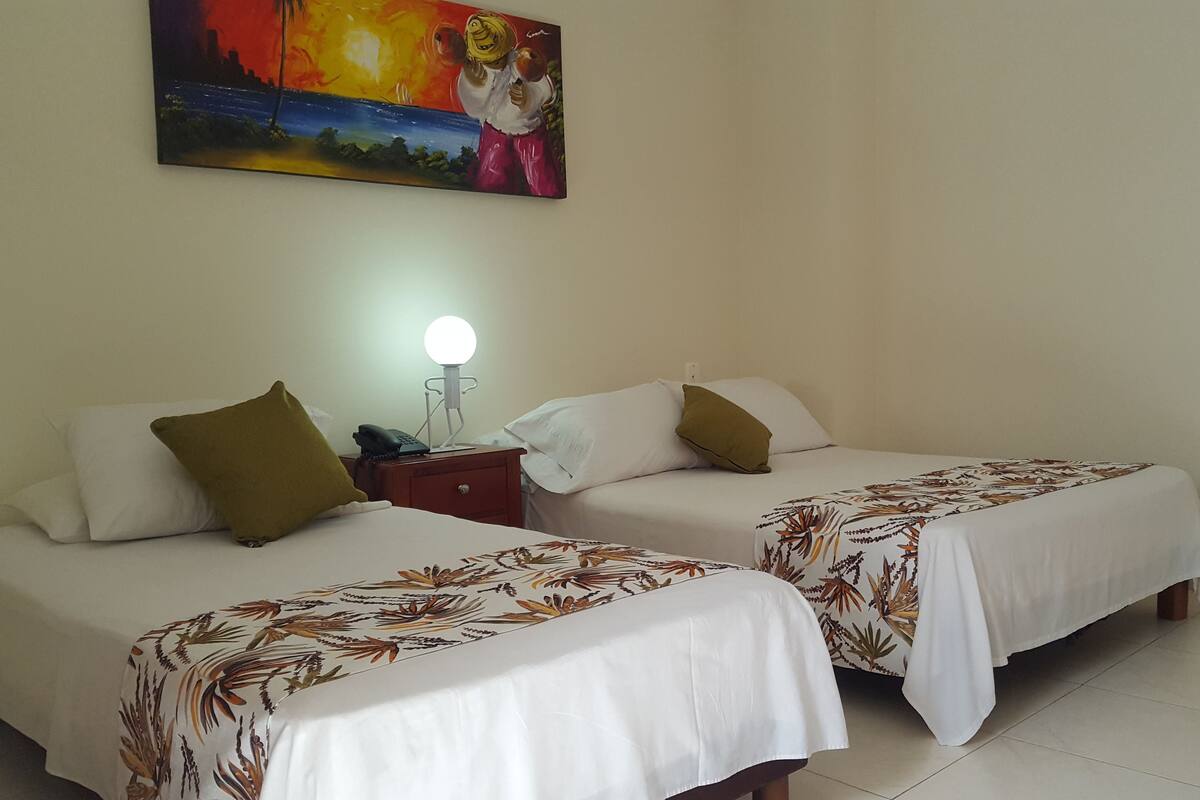 Quarto de hotel com duas camas, mesa de cabeceira com abajur, um quadro na parede e um telefone.
