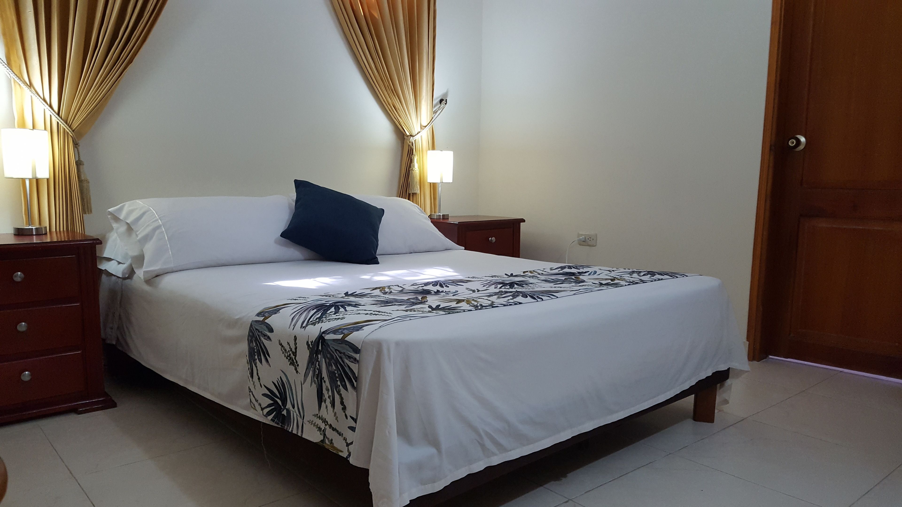 Cama bem arrumada com colcha florida, duas mesas de cabeceira com abajures e uma porta de madeira.