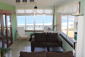 Uma sala de estar com vista para a praia, um sofá, uma poltrona, uma mesinha e uma estante de livros.