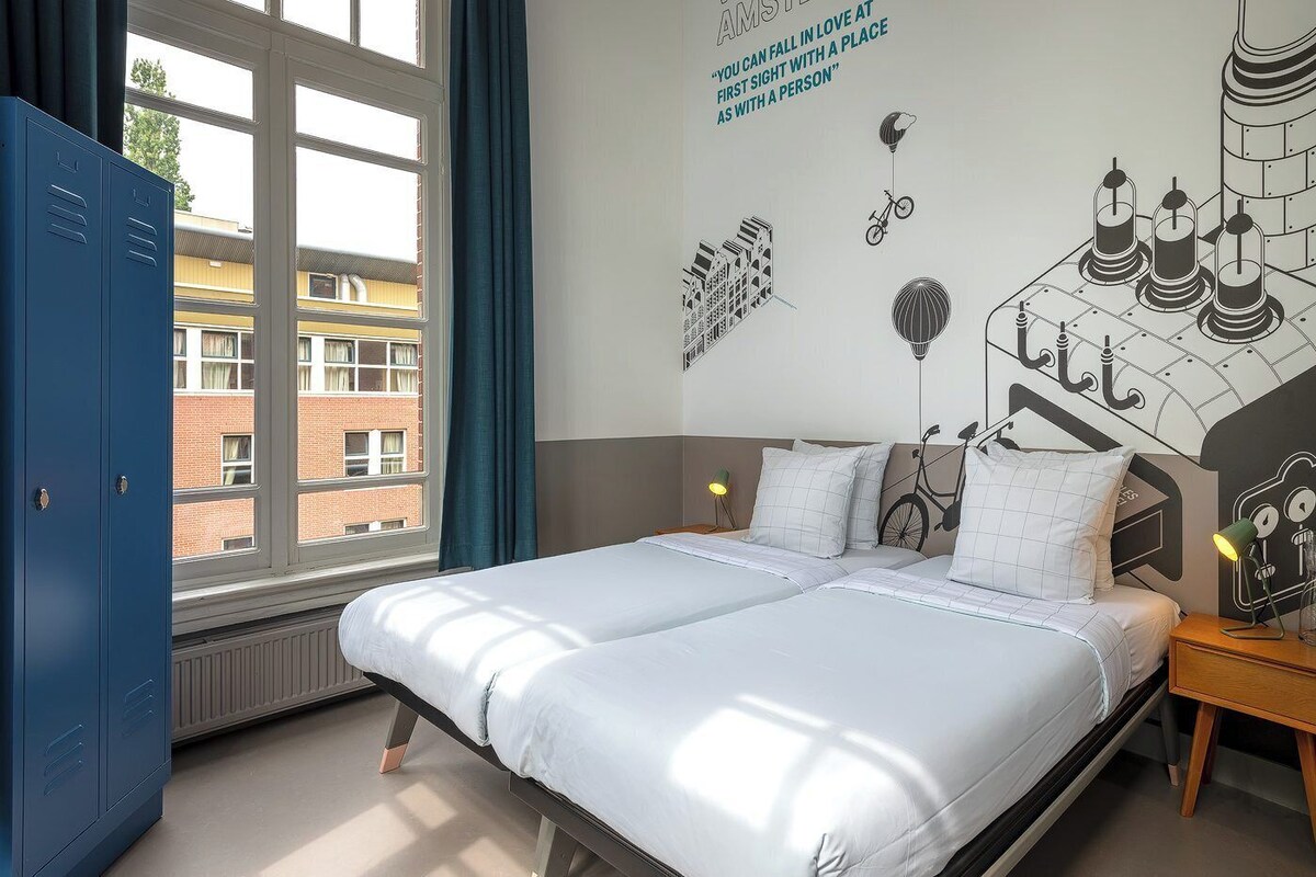 Um quarto com cama, guarda-roupa, janela com cortinas e um mural na parede retratando uma paisagem urbana.