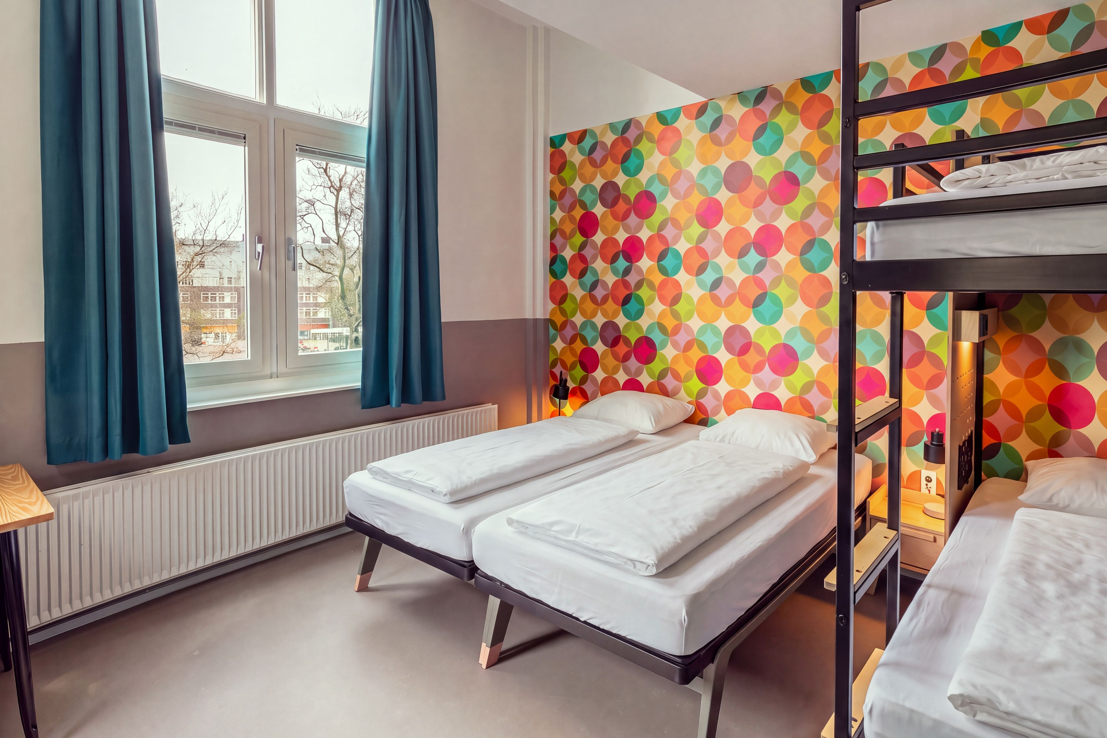 Quarto de hotel com duas camas, uma janela com cortinas, um radiador e uma parede colorida.