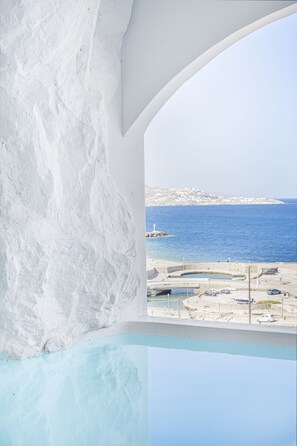 Vista de um quarto com parede branca e janela para área costeira, piscina e cais.