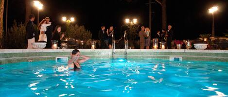 Uma piscina com pessoas ao redor, cercada por árvores e postes de luz.