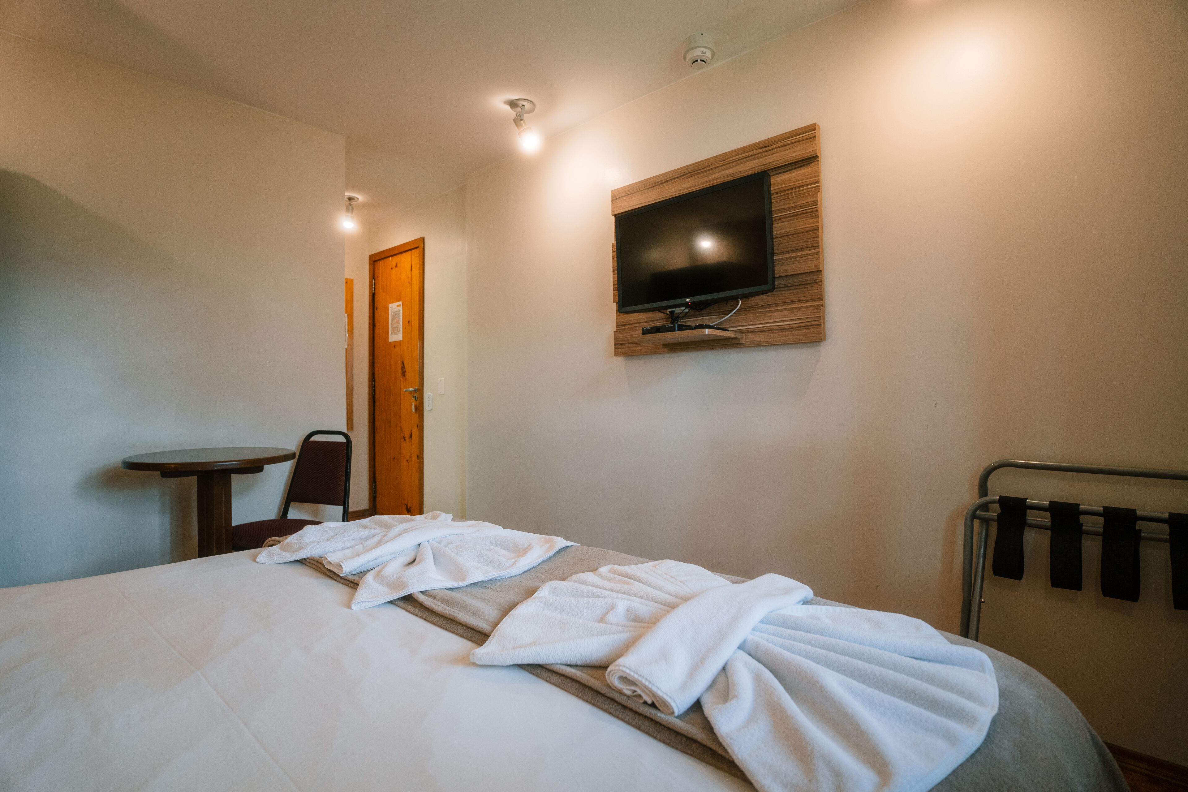 Quarto de hotel com uma cama, televisão fixada na parede, mesa com uma cadeira e uma porta.