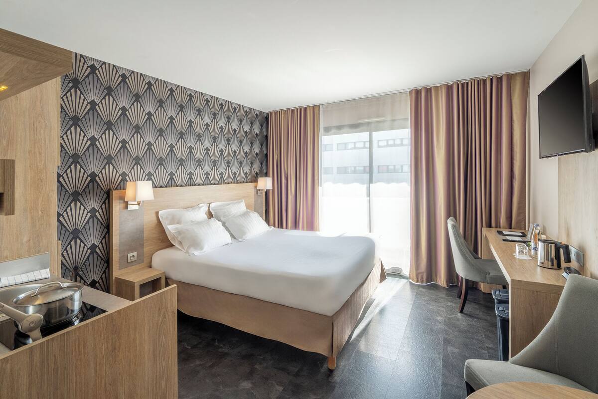 Quarto de hotel moderno com uma cama grande, uma escrivaninha com cadeira, uma televisão de tela plana e vista para a cidade.