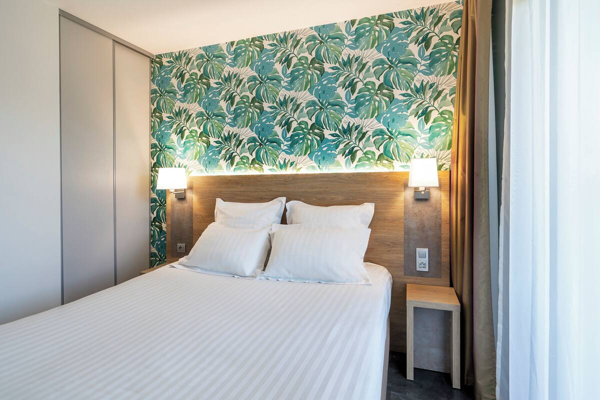 Quarto de hotel com cama, duas luminárias de cabeceira, mesa de apoio e parede com padrão de folhagem tropical.