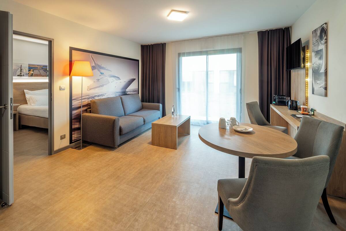 Quarto de hotel moderno com sofá, mesa de jantar redonda, máquina de café e televisão fixada na parede.