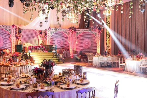 Uma sala de banquetes com mesas redondas preparadas para um evento formal, decorada com arranjos florais e louças elegantes.