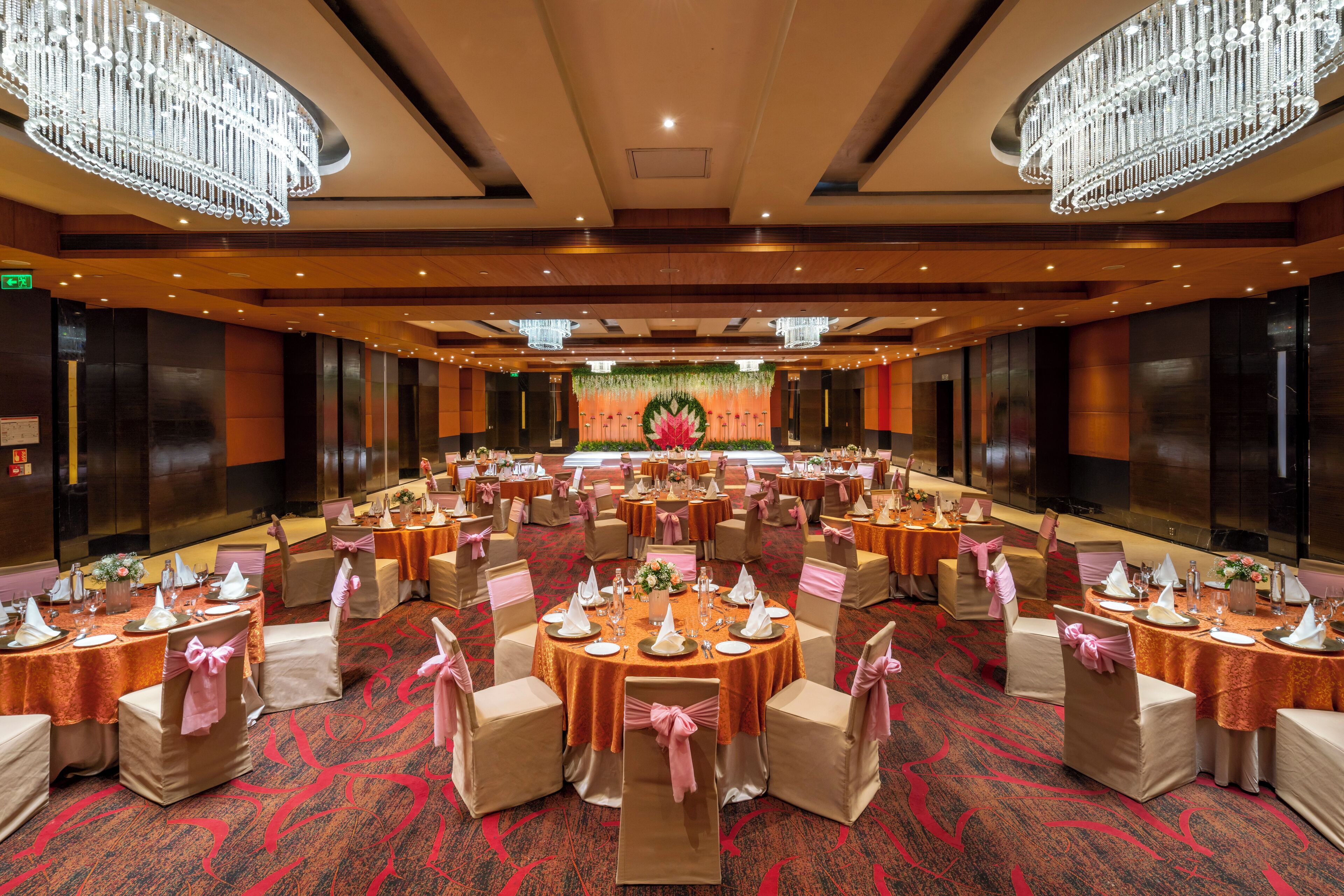 Salão de banquetes com mesas redondas, cadeiras e um painel decorado com flores.
