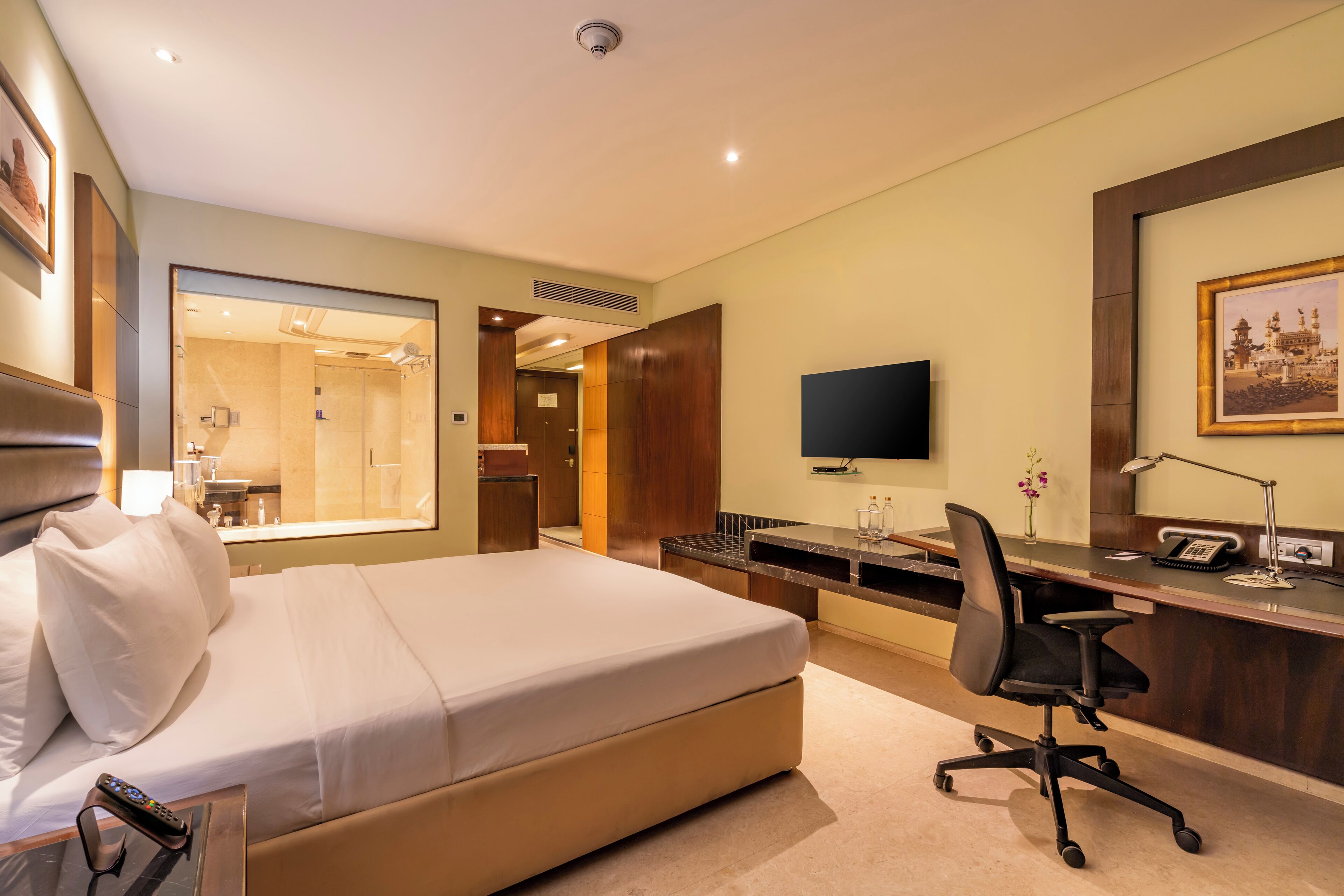 Quarto de hotel com uma cama grande, uma escrivaninha com computador, televisão e banheiro visível através de uma porta aberta.
