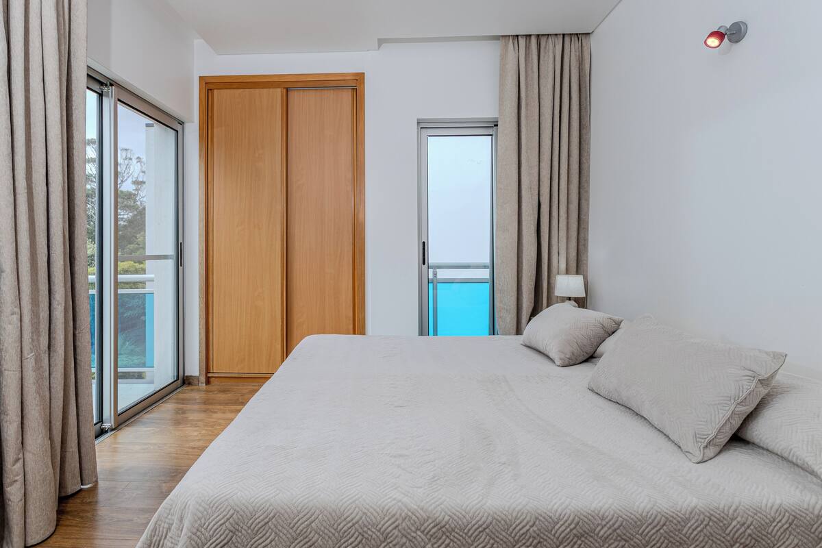 Quarto com cama grande, porta de vidro deslizante, guarda-roupa de madeira e mesa de cabeceira com abajur.