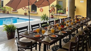 Uma área de refeições com mesas preparadas para o café da manhã, guarda-sóis de exterior e uma piscina ao fundo.