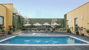 Piscina com espreguiçadeiras e guarda-sóis, rodeada por plantas em vasos e fachada de um edifício amarelo.