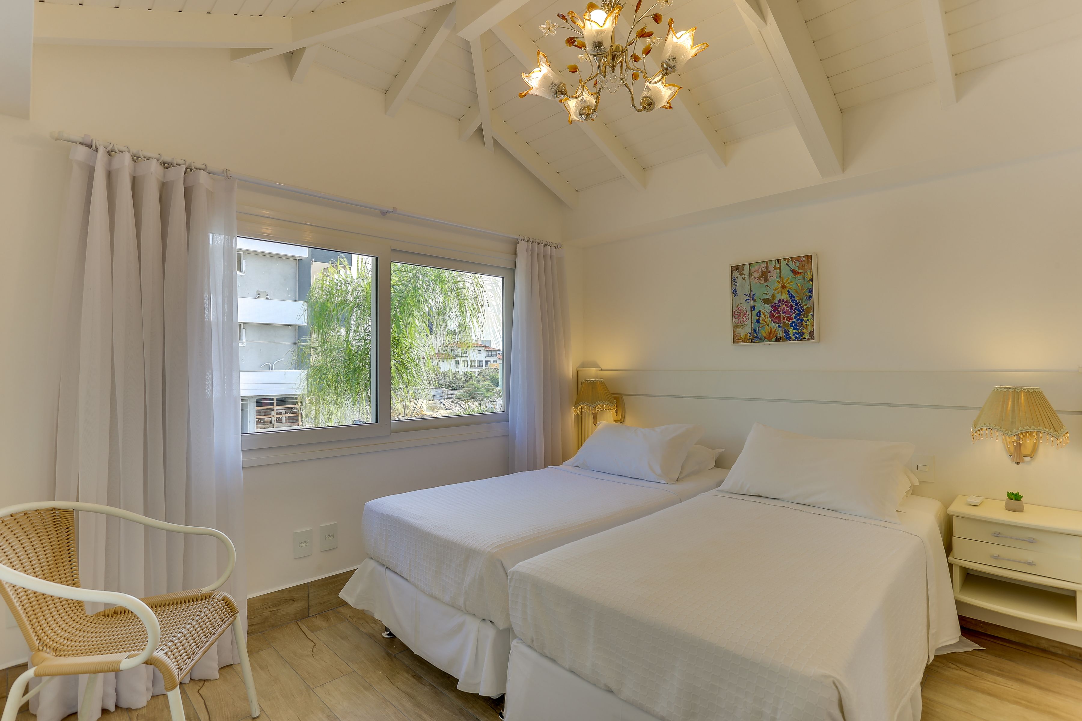 Quarto com duas camas, uma cadeira de vime, janela com cortinas, lustre e mesa de cabeceira com abajur.