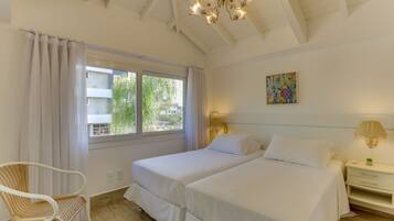 Quarto com duas camas, uma cadeira de vime, janela com cortinas, lustre e mesa de cabeceira com abajur.
