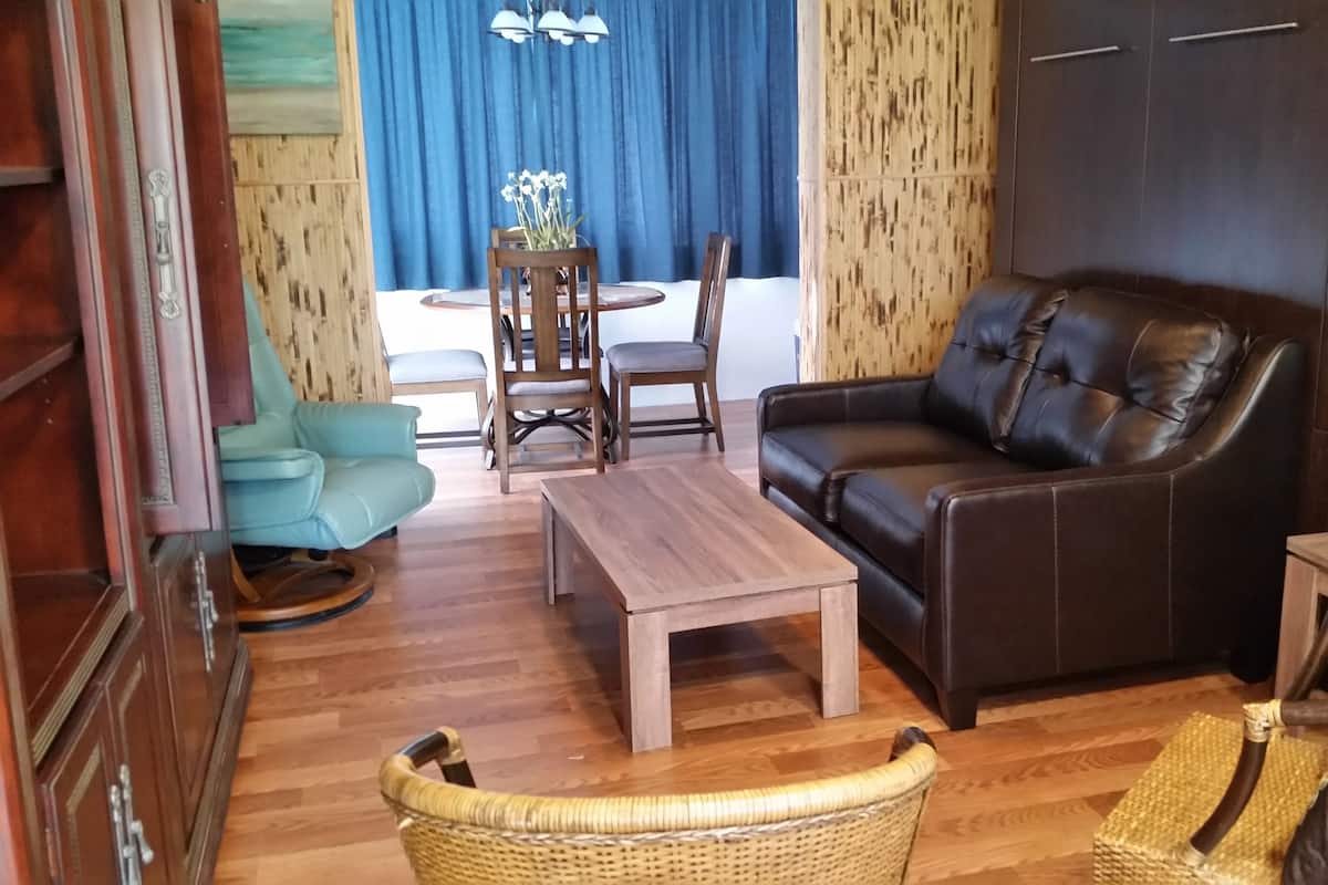 Uma sala de estar com um sofá de couro escuro, mesa de centro de madeira, cadeiras de vime e um espaço de jantar com mesa e cadeiras.