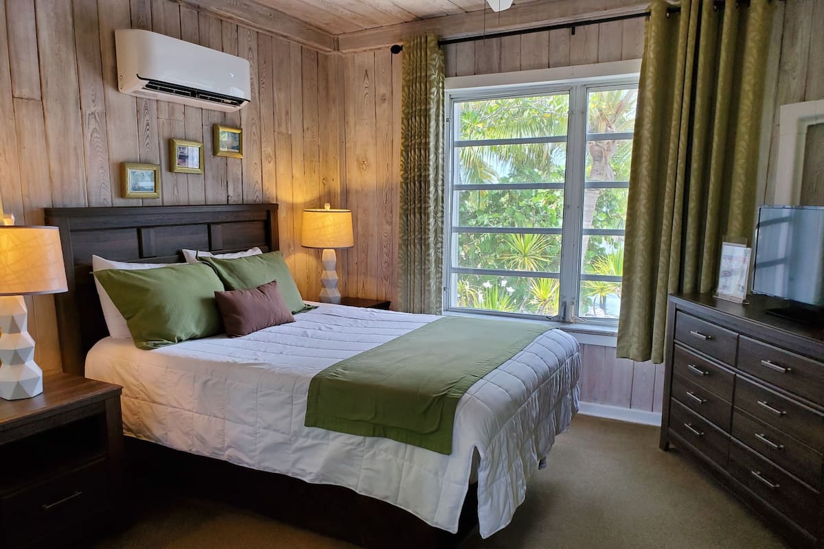 Um quarto com cama, duas mesas de cabeceira com luminárias, cômoda, janela com cortinas e ar condicionado na parede.