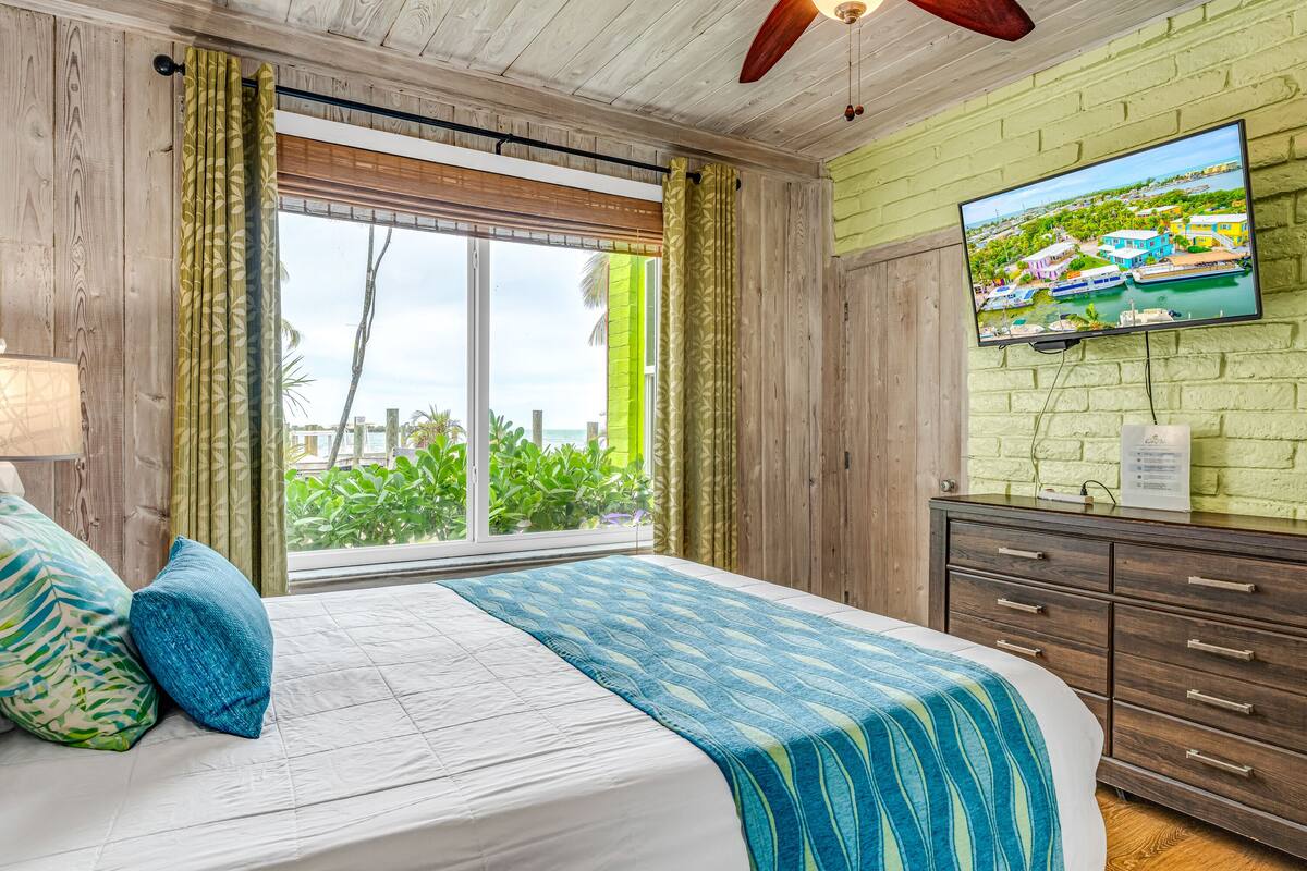 Quarto com cama, televisão na parede, janela com cortinas e cômoda de madeira.