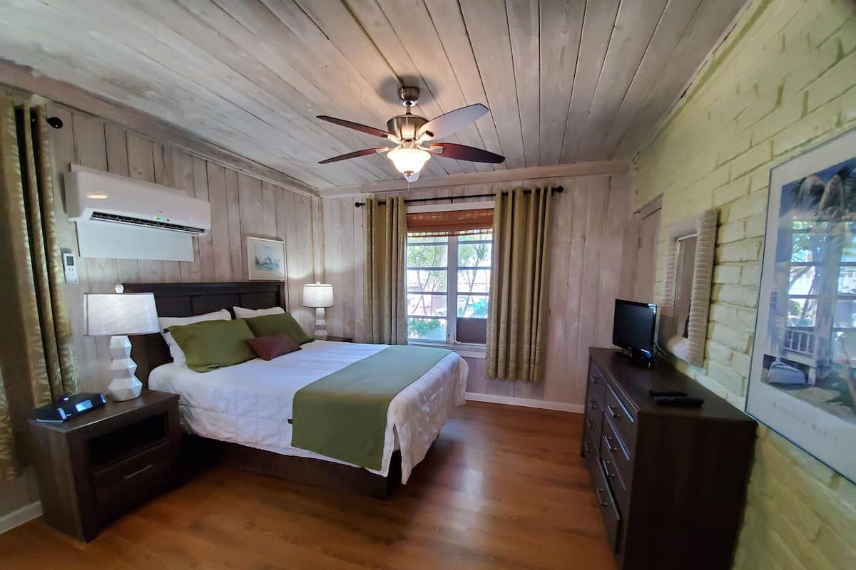 Quarto com painéis de madeira, cama, mesas de cabeceira, ventilador de teto e televisão sobre um aparador.