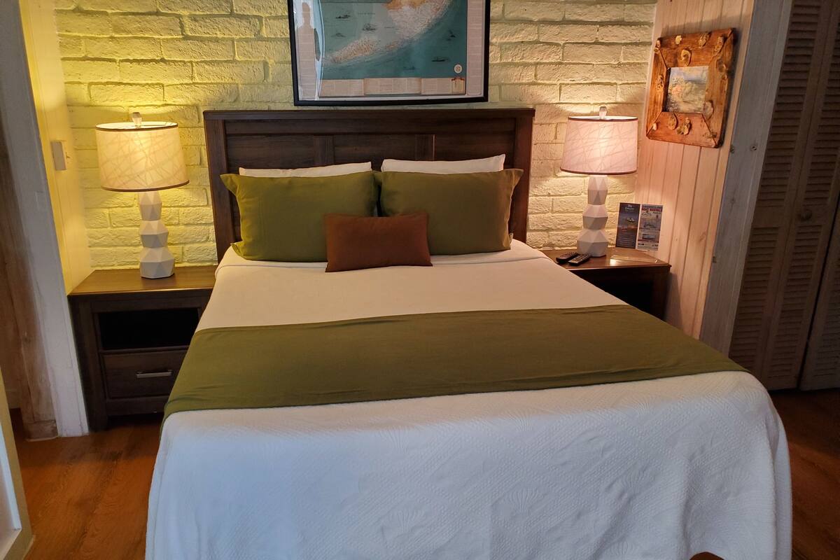 Cama bem arrumada com edredom branco e manta verde, acompanhada por duas mesas de cabeceira com abajures, um mapa emoldurado na parede e um porta-retrato de madeira.
