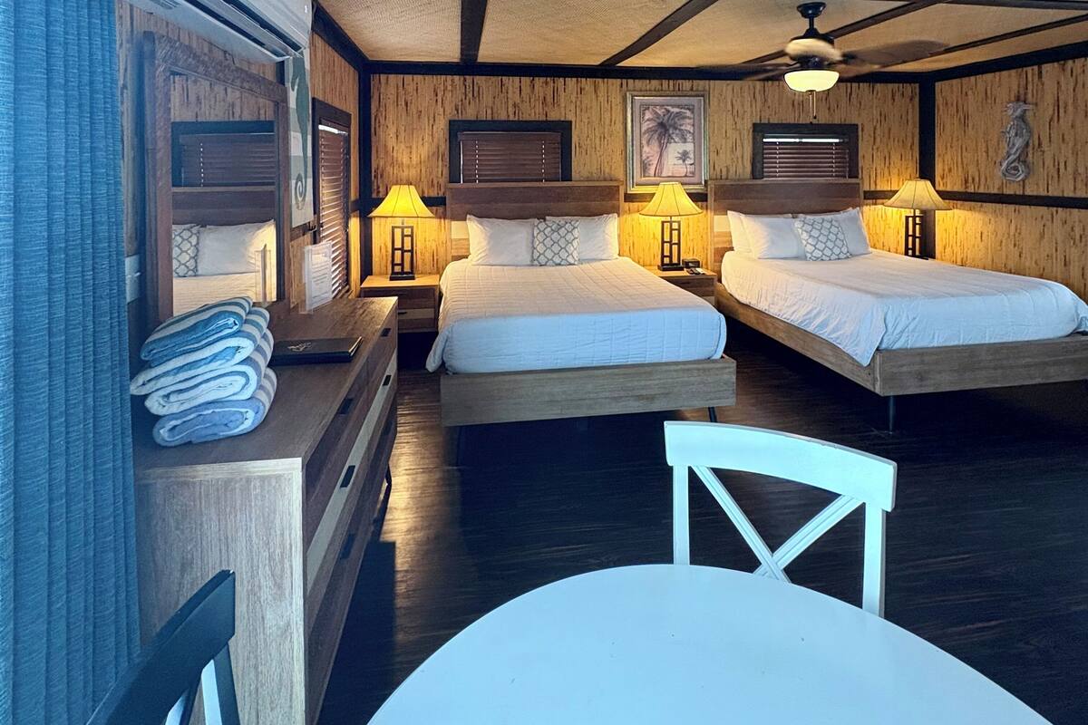 Quarto de hotel com duas camas, uma escrivaninha e uma cadeira. Há um ventilador de teto e uma janela com persianas.