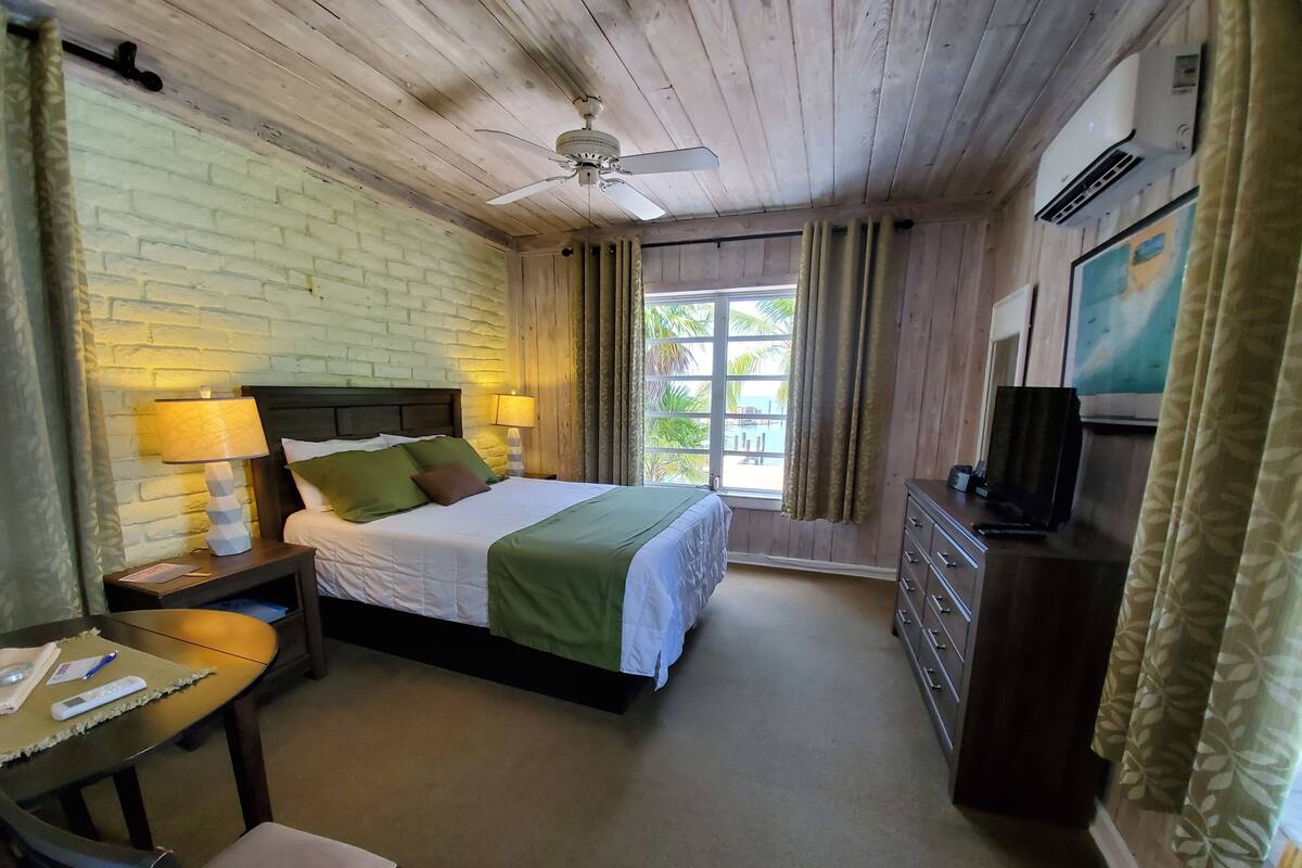 Quarto com cama, mesas de cabeceira, escrivaninha, cadeira, televisão e janela com cortinas.