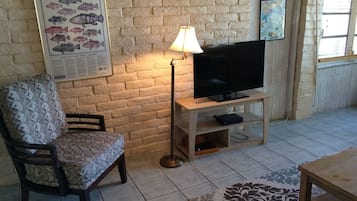 Uma sala de estar com parede de tijolos, televisão sobre um suporte de madeira, poltrona estampada e uma luminária.