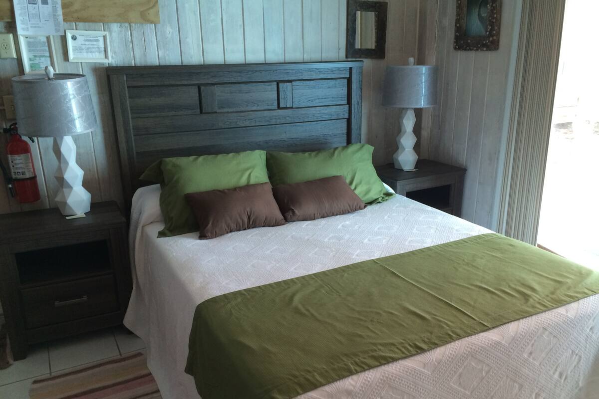 Cama bem arrumada com almofadas verdes e castanhas, cabeceira de madeira e colcha branca. Há duas mesas de cabeceira com luminárias e um extintor de incêndio à esquerda.
