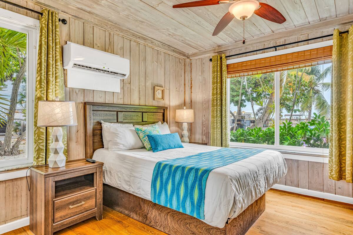 Quarto com teto de madeira, cama com cobertor listrado azul e branco, duas luminárias de cabeceira, mesa de cabeceira com gavetas e janela com cortinas.