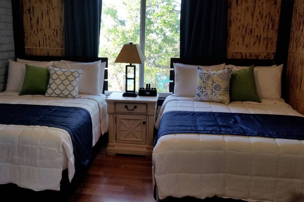 Quarto duplo com duas camas, cada uma com edredom branco, almofadas verdes e estampadas, mesa de cabeceira com abajur e uma gavetinha.