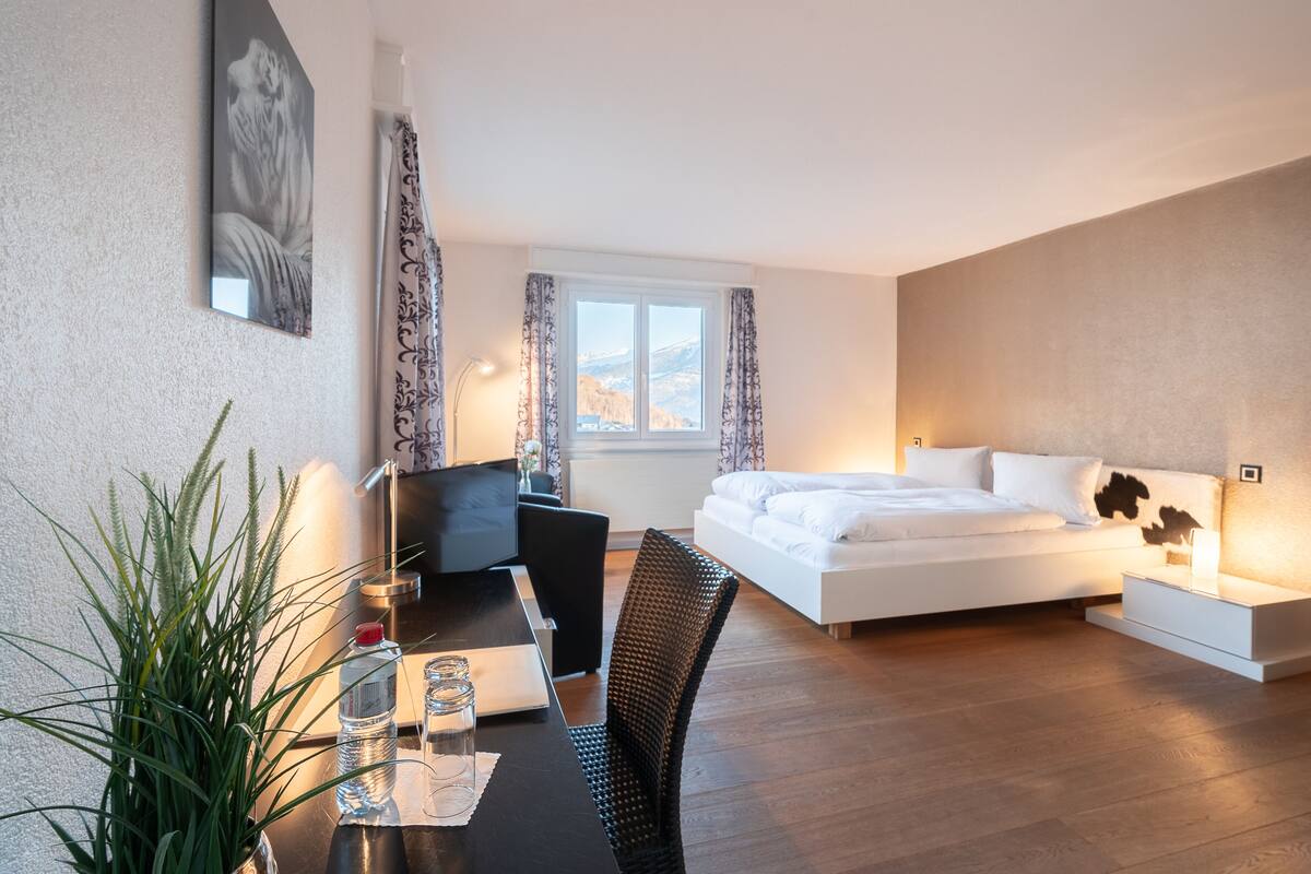 Quarto de hotel moderno com cama, escrivaninha com computador, cadeira, planta em vaso e janela com cortinas.
