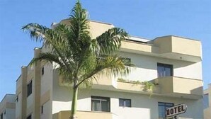 Um hotel com vários andares, com uma varanda no segundo andar, uma palmeira na frente e uma placa indicando ser um hotel.