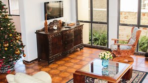 Uma sala de estar com árvore de Natal, televisão, armário de madeira, sofá, mesa de centro e vista através de grandes janelas.