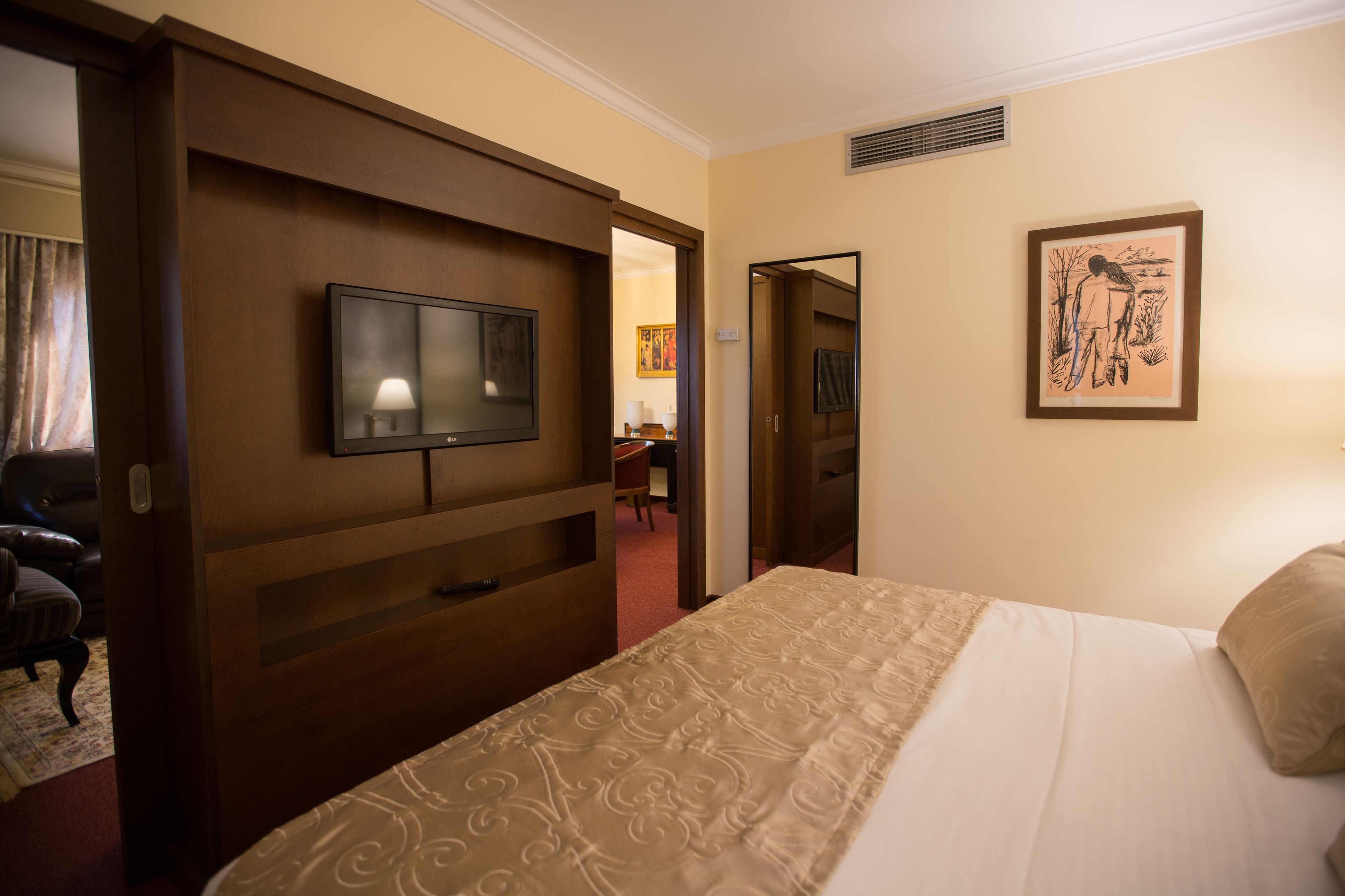 Quarto de hotel com uma cama, televisão fixada em um painel de madeira, uma obra de arte emoldurada na parede e uma porta que dá para outro cômodo.