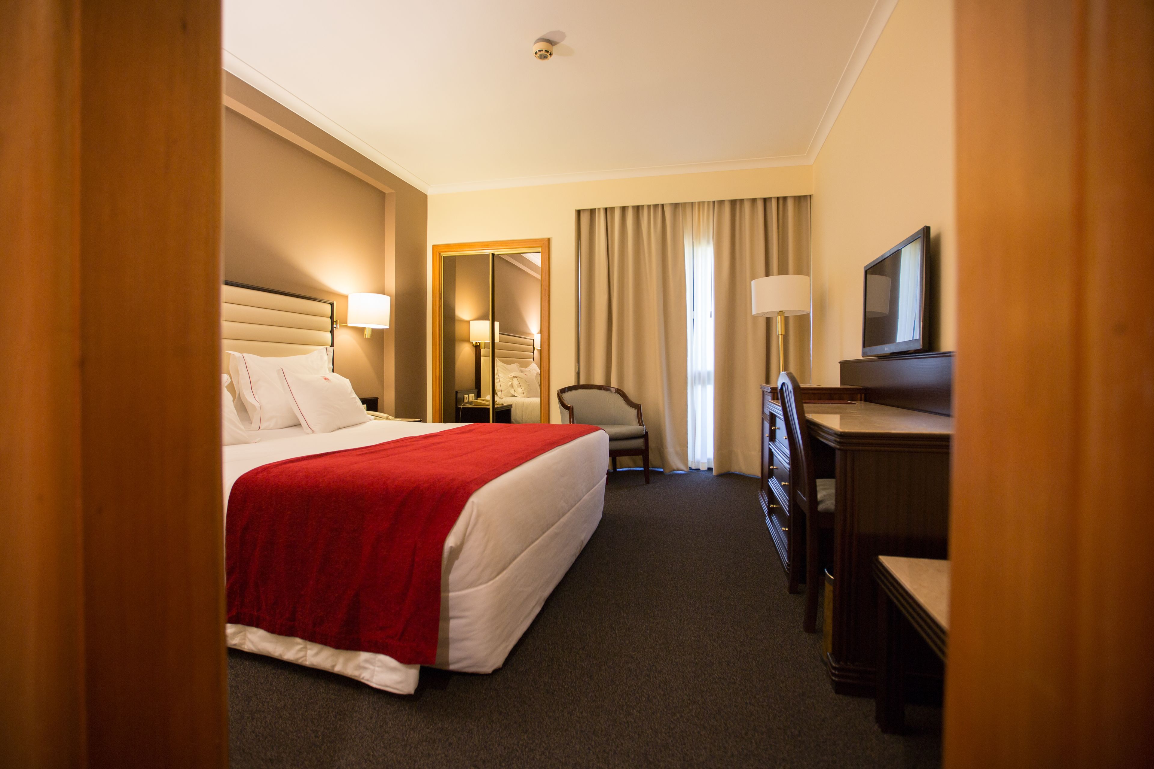 Quarto de hotel com uma cama grande, uma escrivaninha, uma cadeira, uma televisão e um espelho.