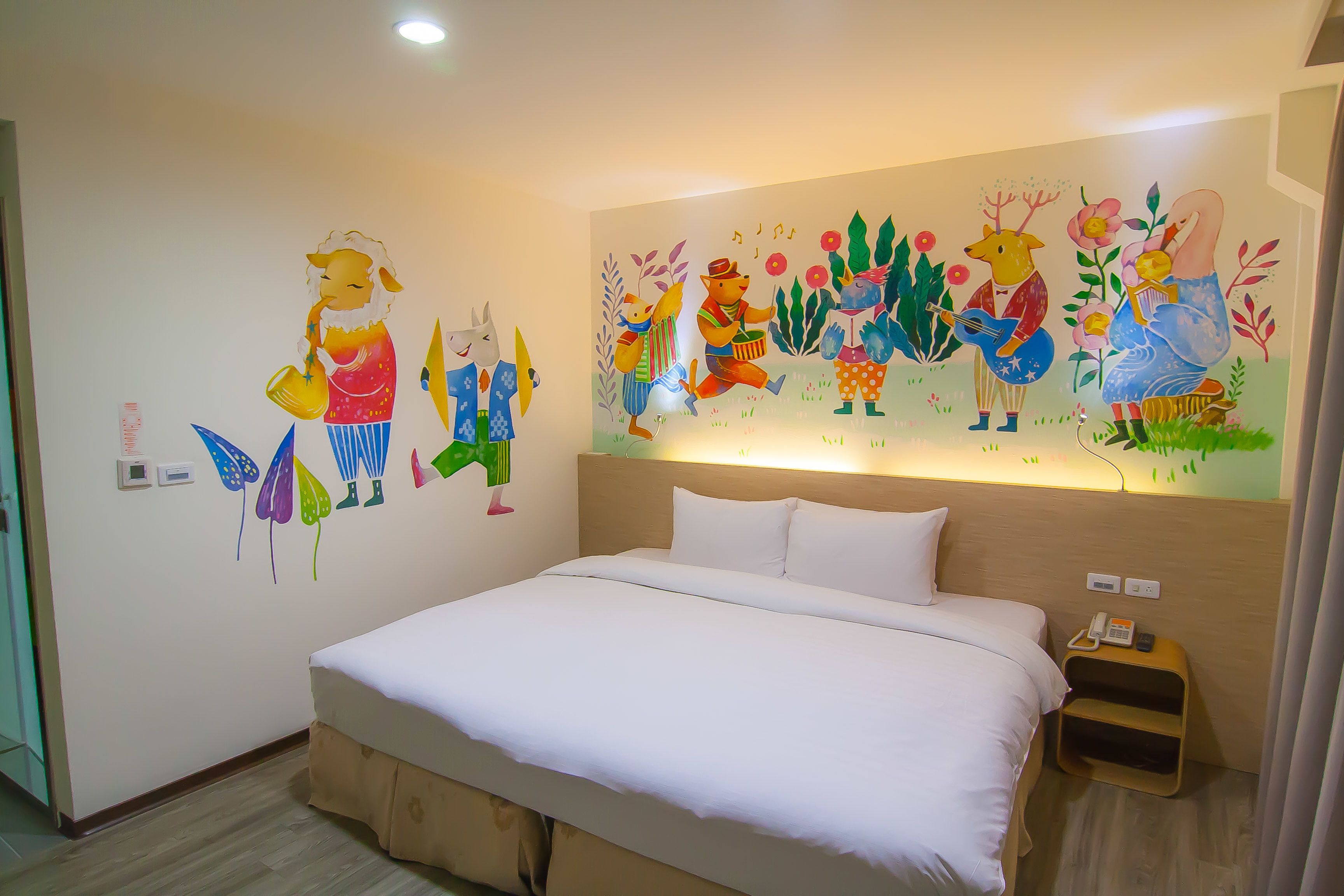 Quarto de hotel com uma cama grande, uma mesinha de cabeceira e um mural na parede retratando animais com características humanas tocando instrumentos musicais.