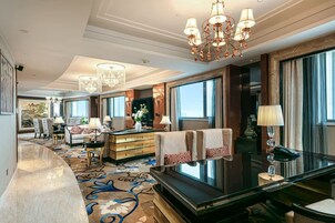 Quarto de hotel espaçoso, com uma cama grande, área de estar com sofá e poltronas, mesa de jantar e uma janela ampla com vista para a cidade.