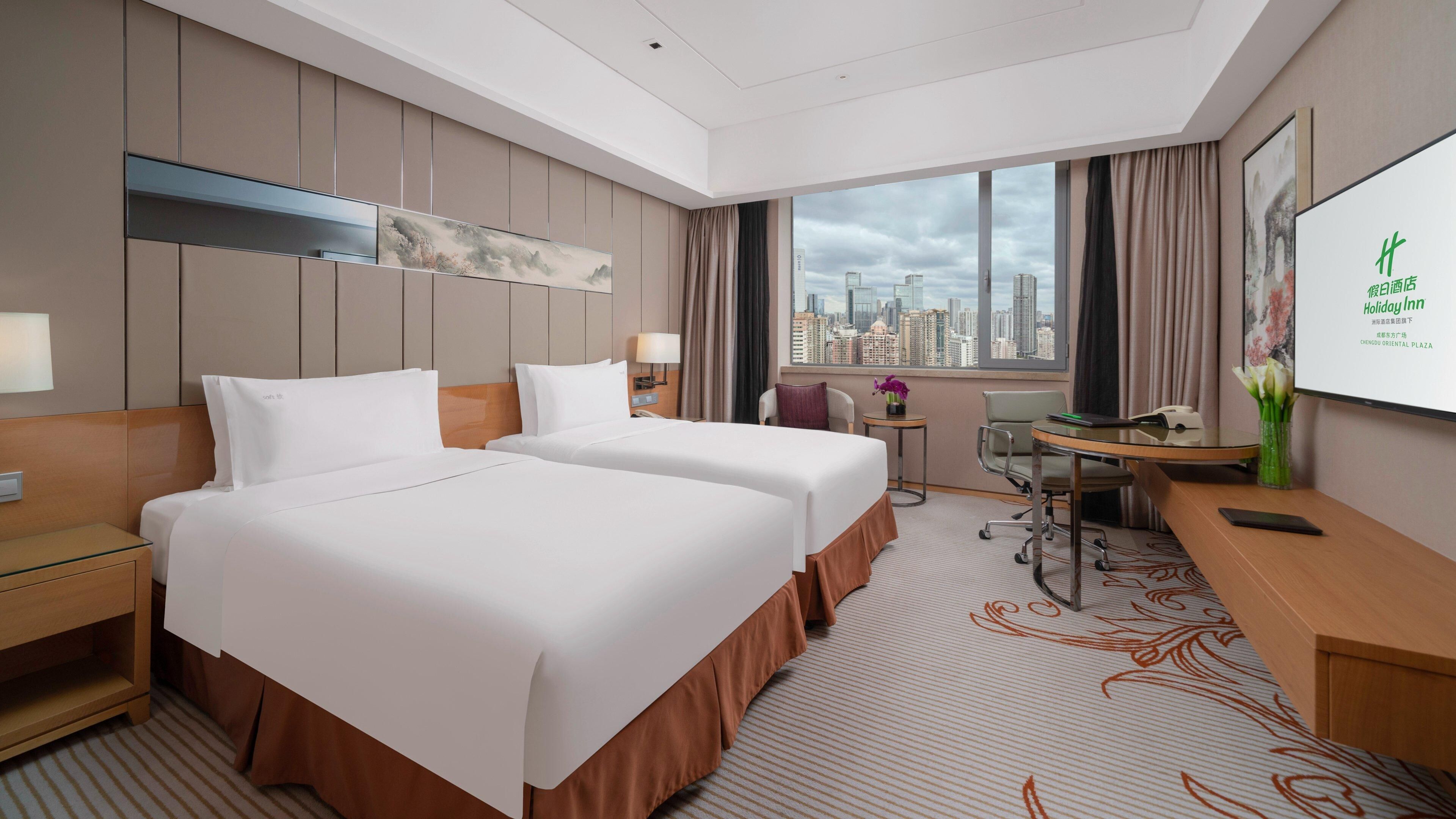 Quarto de hotel com duas camas, uma escrivaninha, uma cadeira, uma mesinha, uma lâmpada, um vaso com flores e vista para a cidade pela janela.