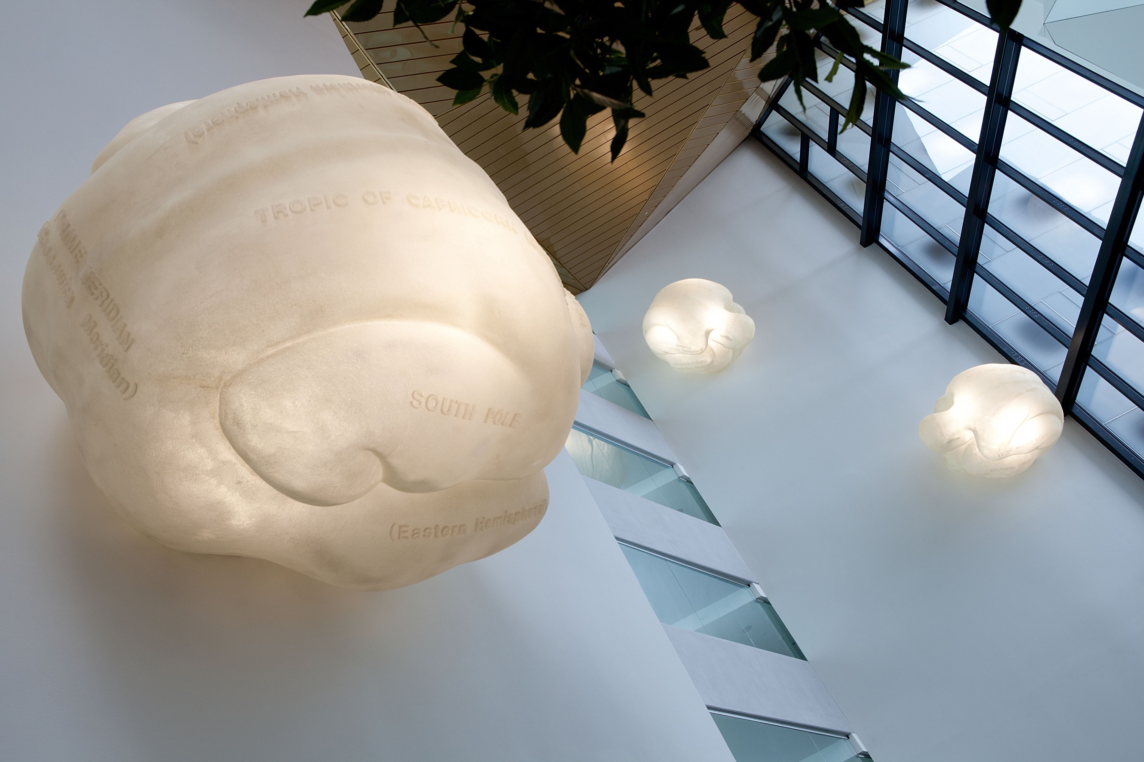 Escultura grande e abstrata com inscrições, guarda-corpo de vidro e uma planta em espaço interno moderno.