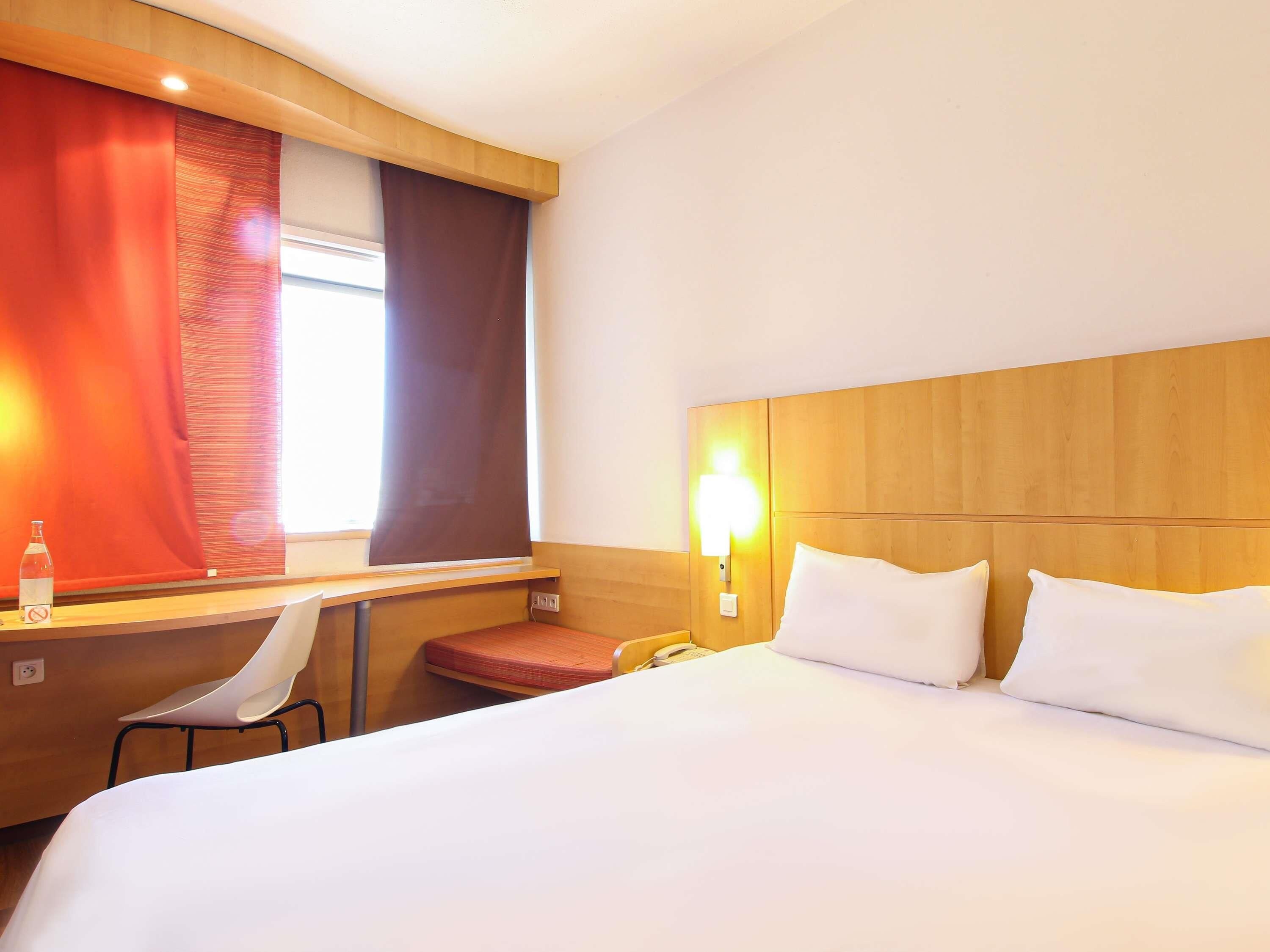 Quarto de hotel com cama, escrivaninha, cadeira, telefone e janela com cortinas.