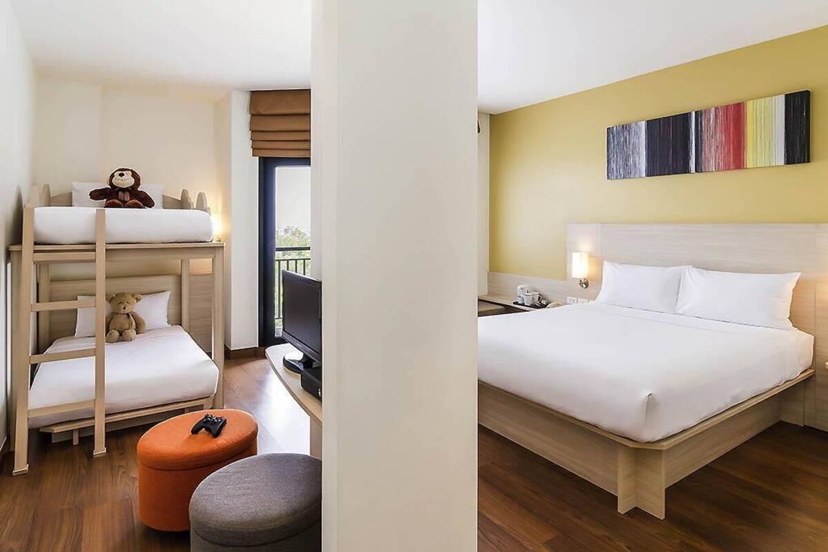 Quarto de hotel com beliche, uma cama de solteiro, televisão e um quadro abstrato colorido na parede.