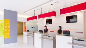 Uma área de recepção de hotel moderna, com três balcões de tampo de vidro, uma mulher atrás de um deles, e uma TV fixada na parede.