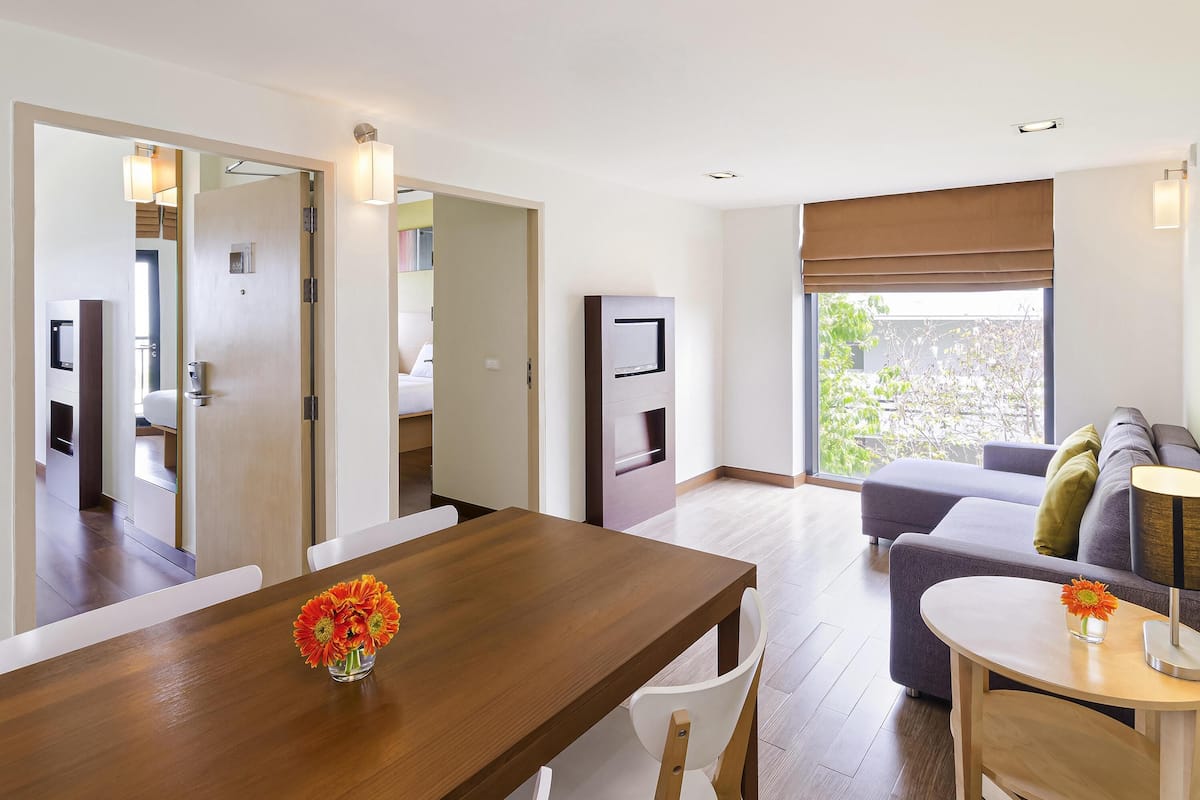 Quarto de hotel moderno com uma mesa de jantar de madeira, cadeiras brancas, um sofá e uma pequena mesa redonda com uma luminária.