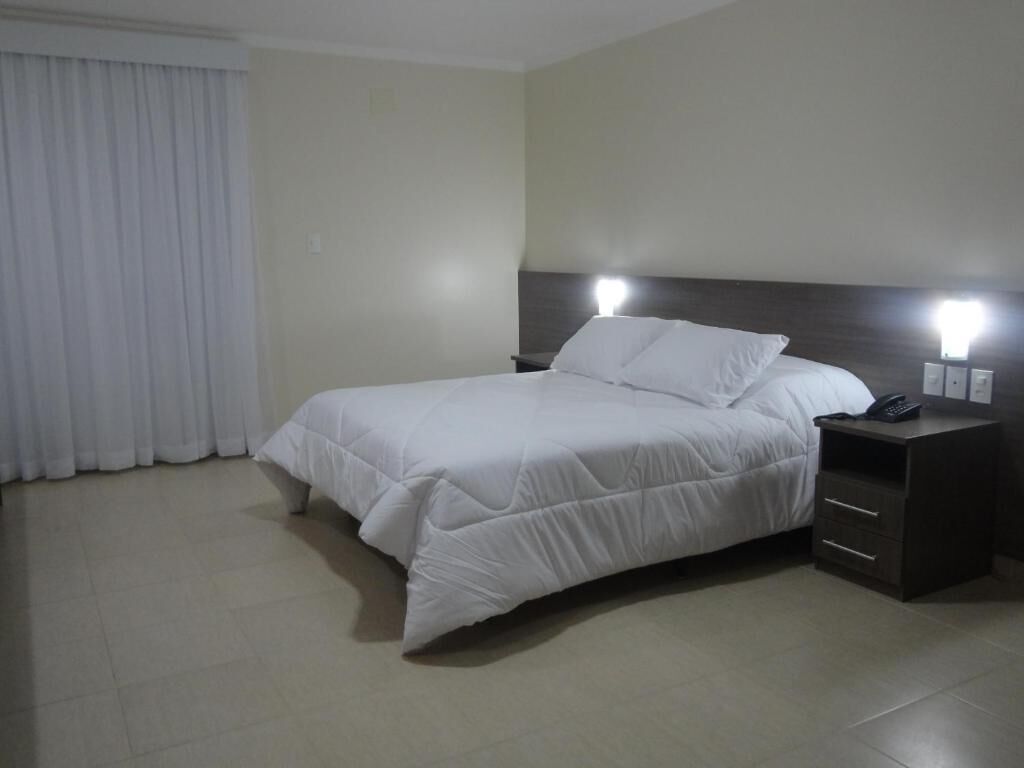 Cama bem arrumada num quarto de hotel, com cabeceira, mesa de cabeceira com abajur e janela com cortinas.