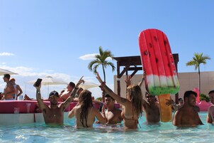 Piscina com pessoas relaxando, cercada por palmeiras e guarda-sóis.